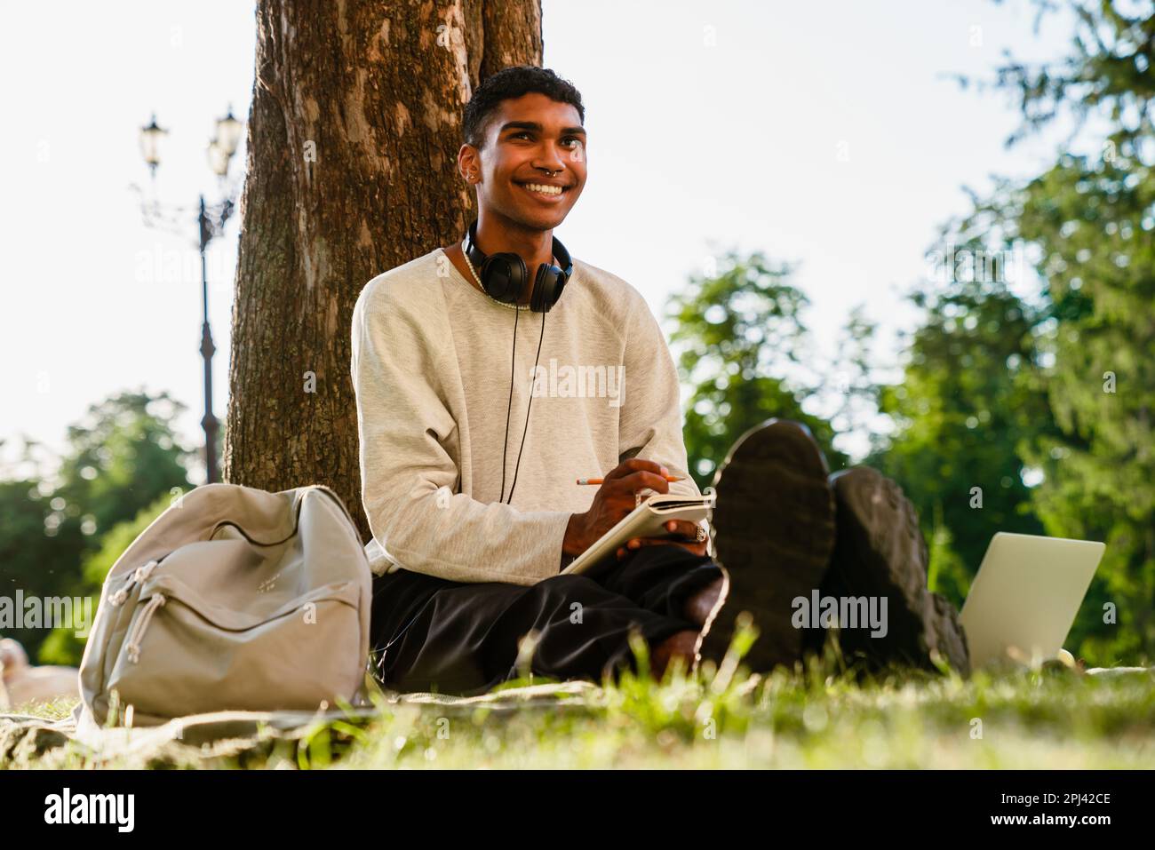 Young black man studying and taking notes while sitting under tree in ...
