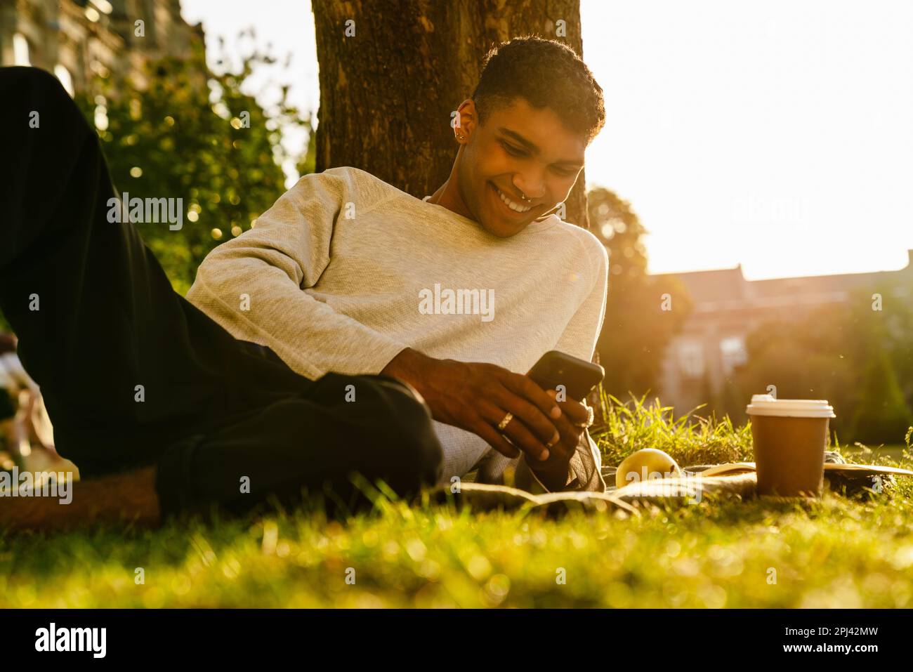 Young black man smiling and using mobile phone while lying under tree ...