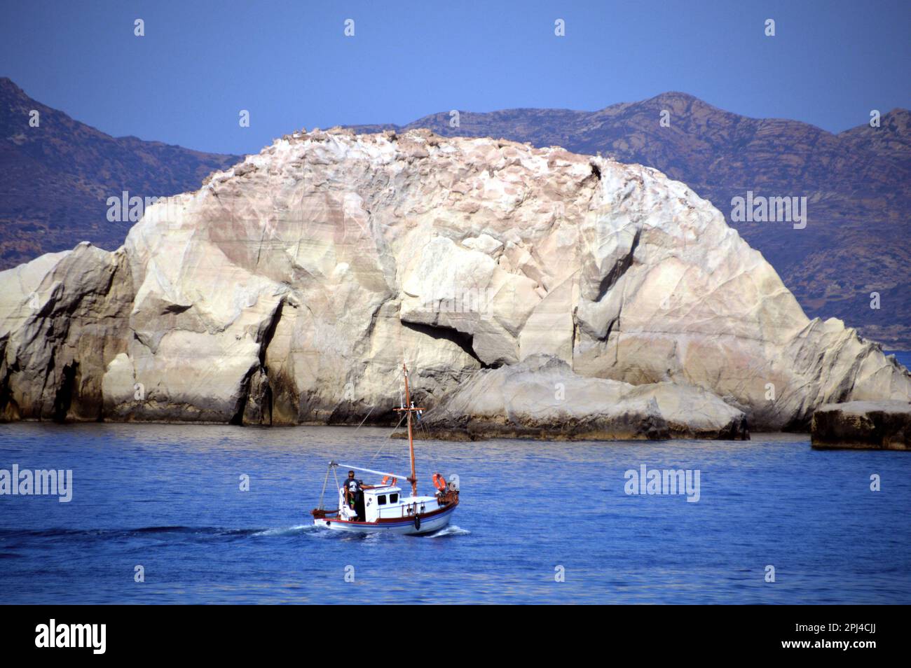 Greece, Island of Milos: fishing boat returning to Mandrakia, dwarfed ...