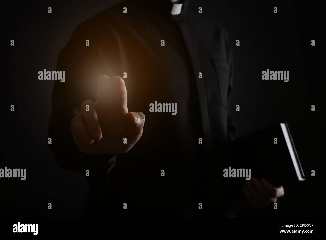 Priest with Bible making blessing gesture and holy light on dark ...