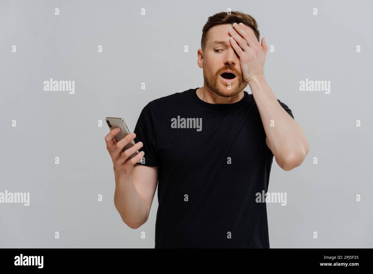 Scared man covers half of face with hand stares amazed at smartphone ...