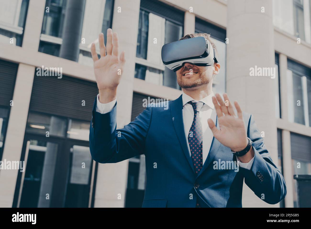 Young cheerful businessman with beard dressed in blue formal suit trying out VR glasses ...