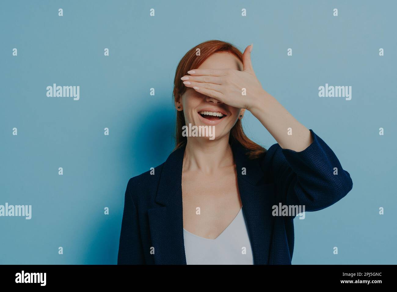 Playful funny ginger girl covering her eyes with palm isolated on blue ...