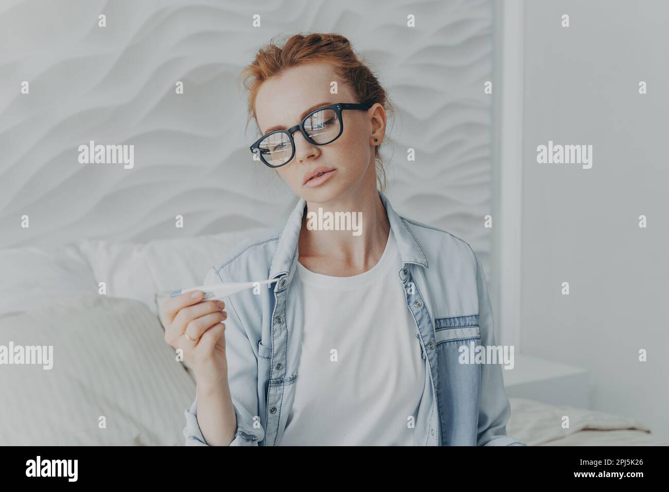 Young unhealthy red-haired lady holding thermometer while sitting on ...