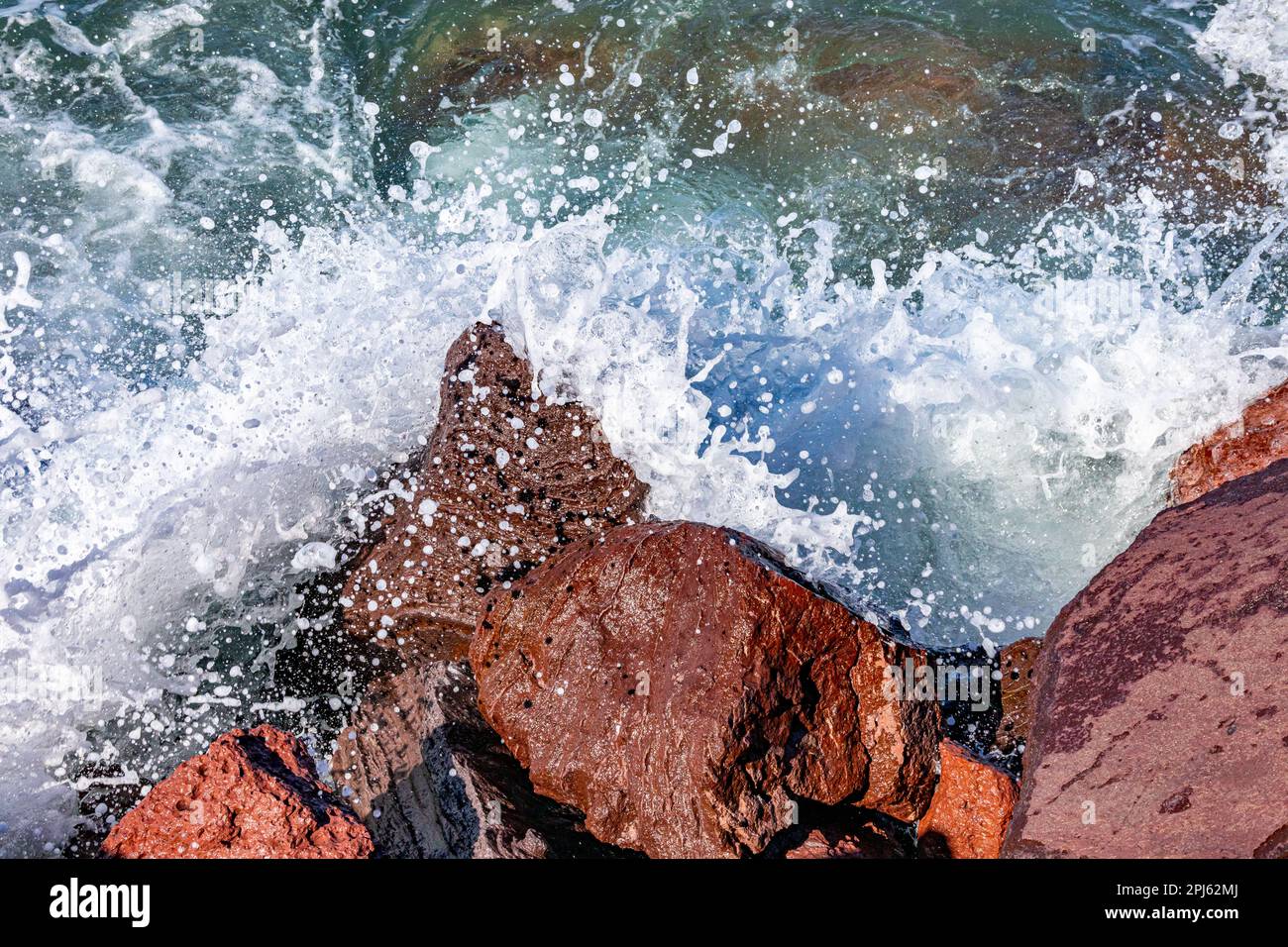 Close up of sea water breaking over reddish brown rocks forming foam ...