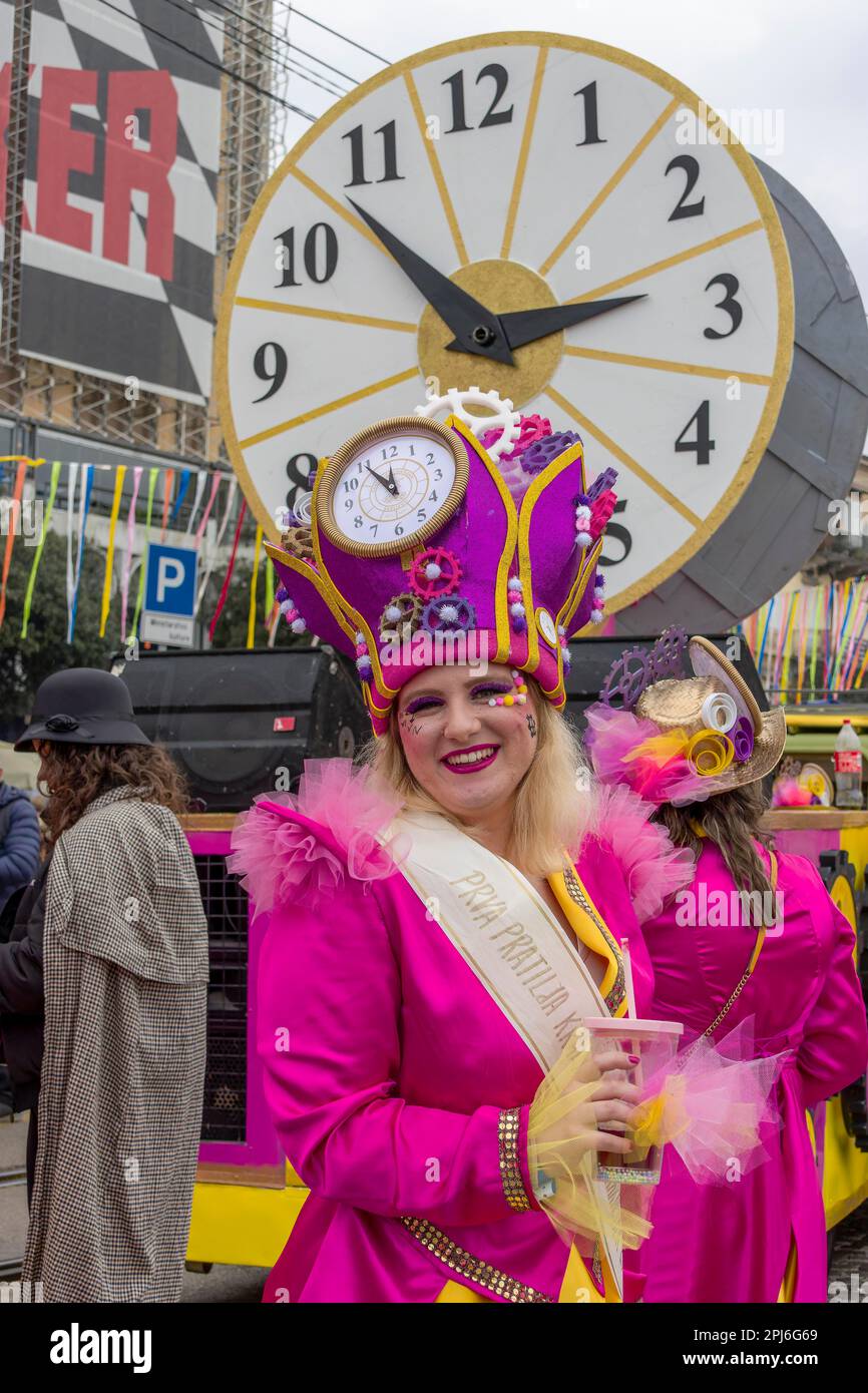 A masked, smiling girl in pink clothes with a clock hat and a big clock ...