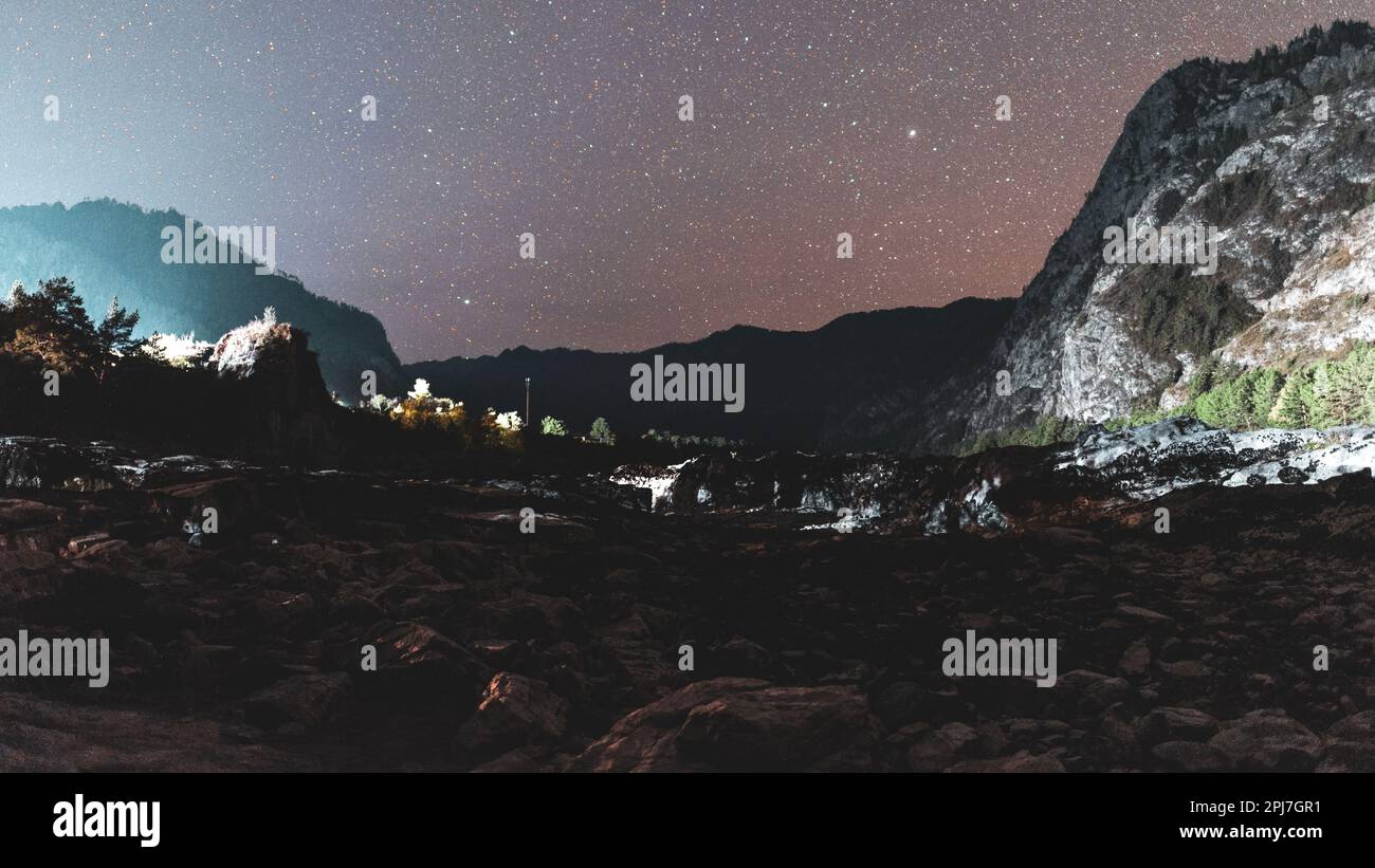 The stone bank of the Katun River in the shadow at night by a light-lit ...