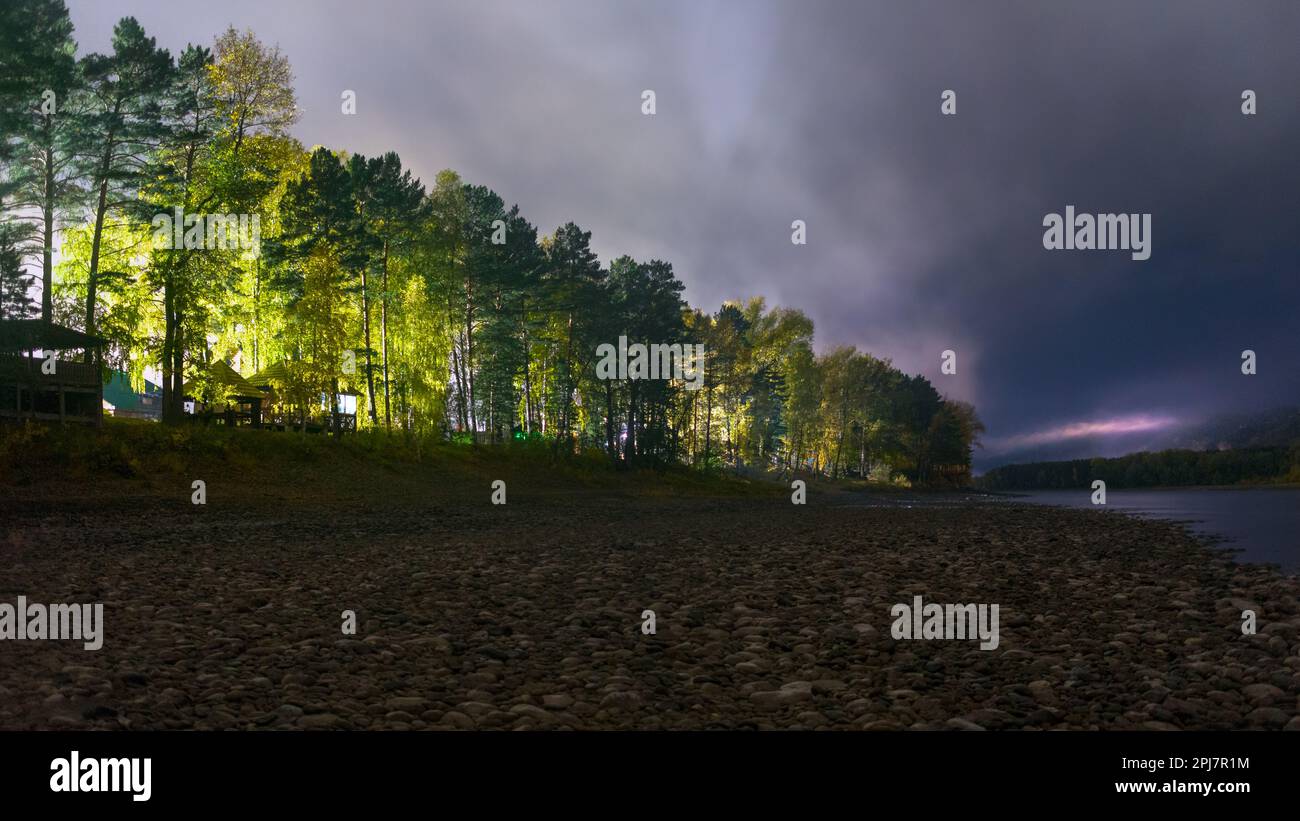 Mountain stone bank of the Katun River with trees at night illuminated ...