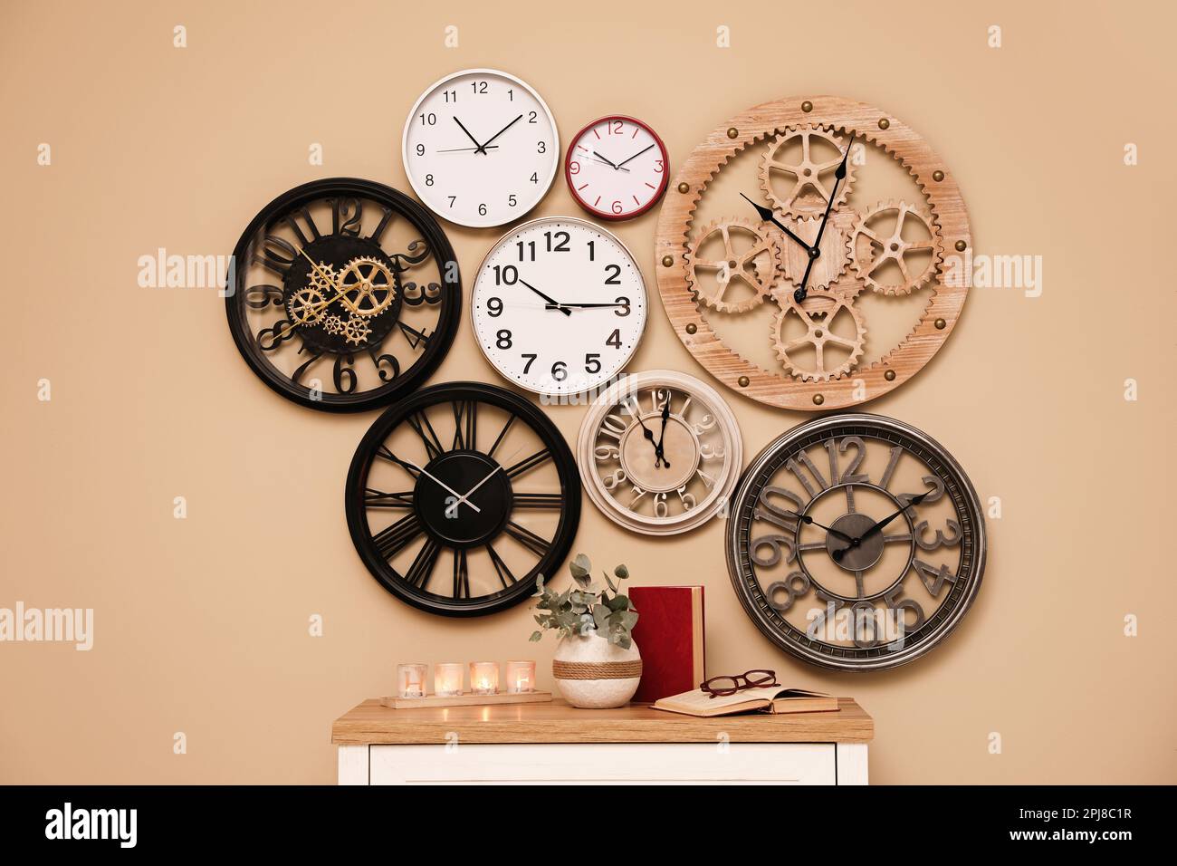 Collection of wall clocks over table with books, candles and plant ...