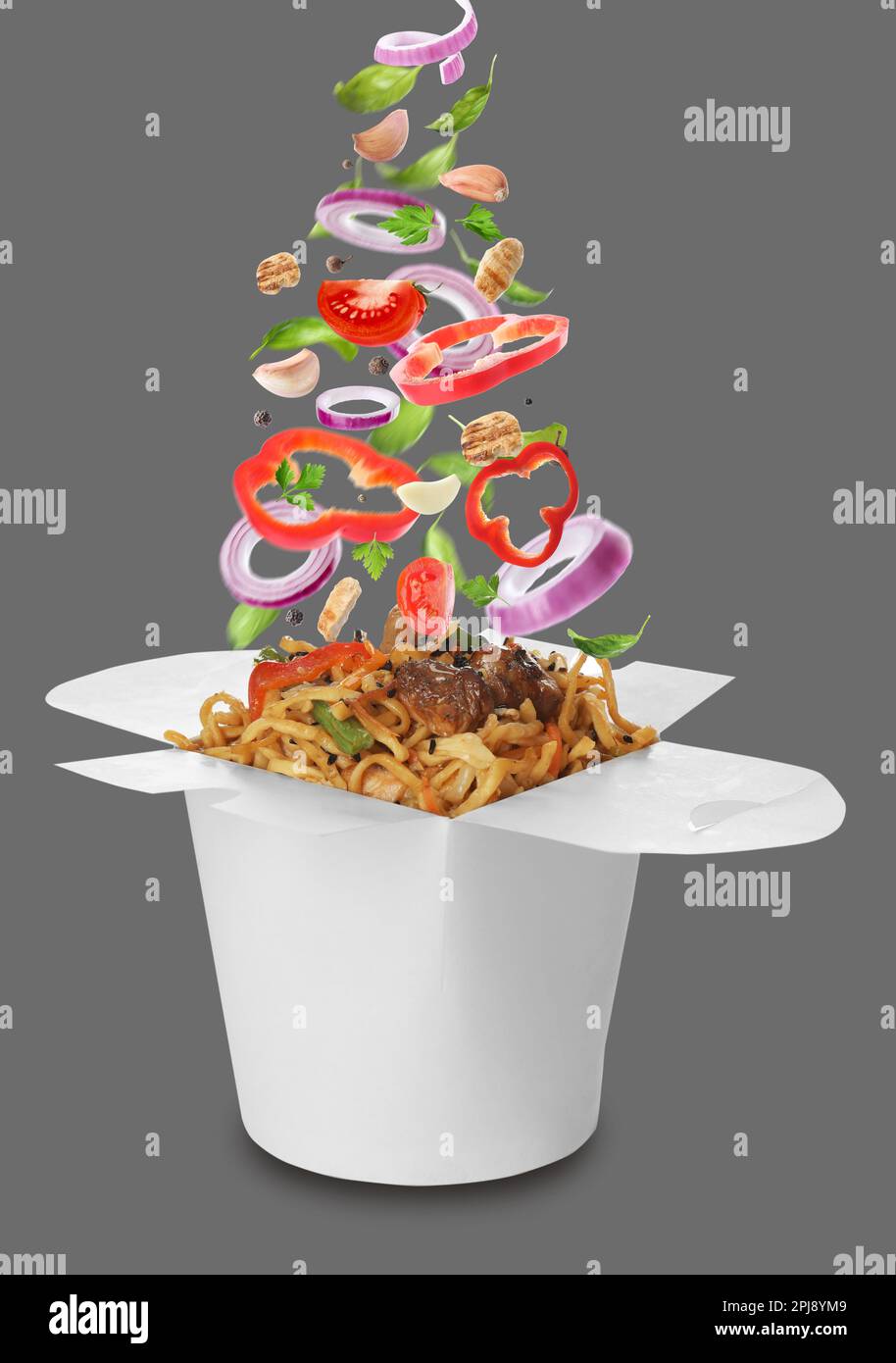 DIfferent fresh ingredients falling into box of wok noodles on grey ...