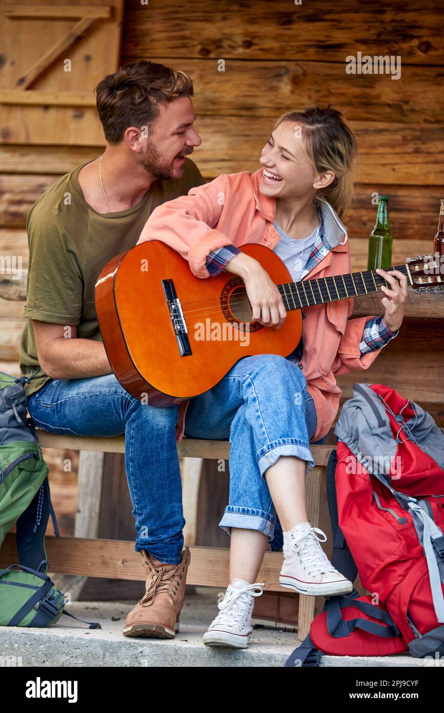 Smiling couple drinking and playing guitar in front of wooden cottage ...