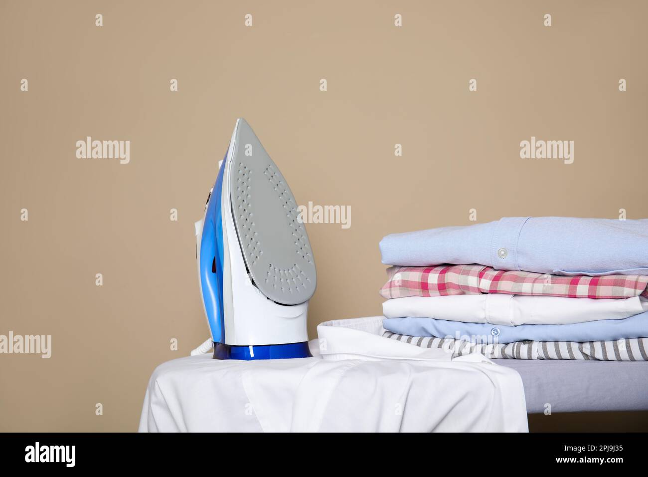 Modern iron, clean shirt and folded clothes on board against beige ...