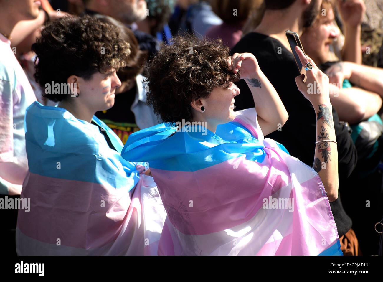 Two people wrapped in the transgender flag listen to speeches during ...