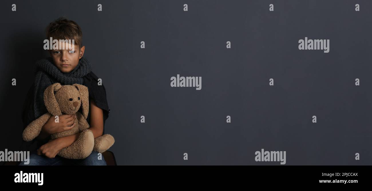 Poor boy sitting with toy rabbit on dark background, space for text ...