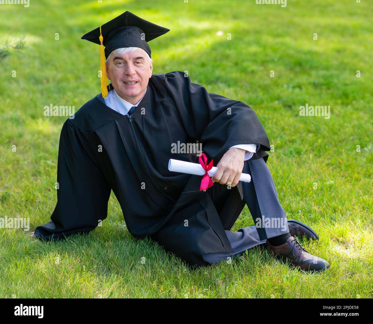 An elderly gray-haired man in a graduation gown sits on green grass ...