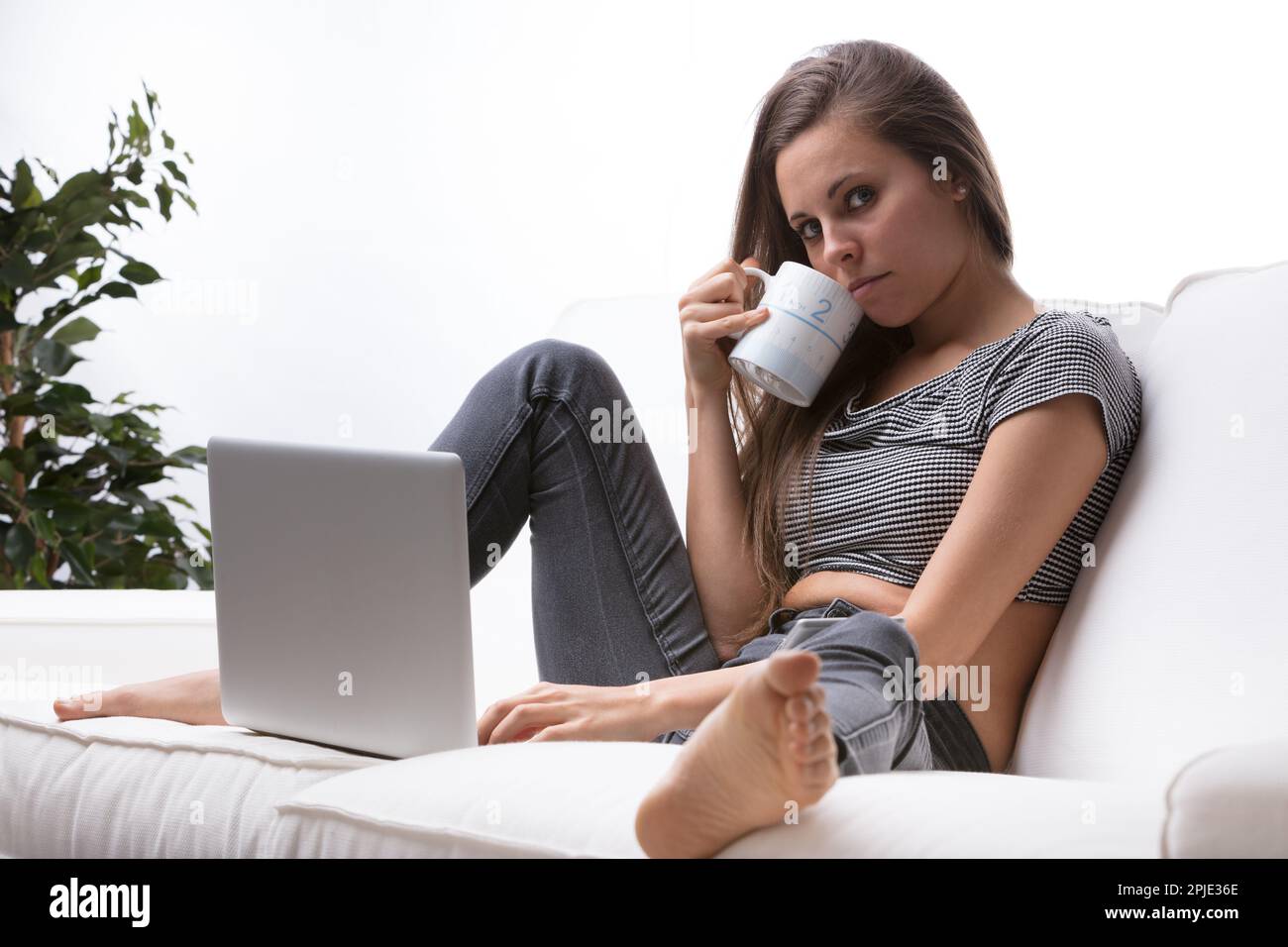 A young woman is sitting on the couch drinking something warm from her ...