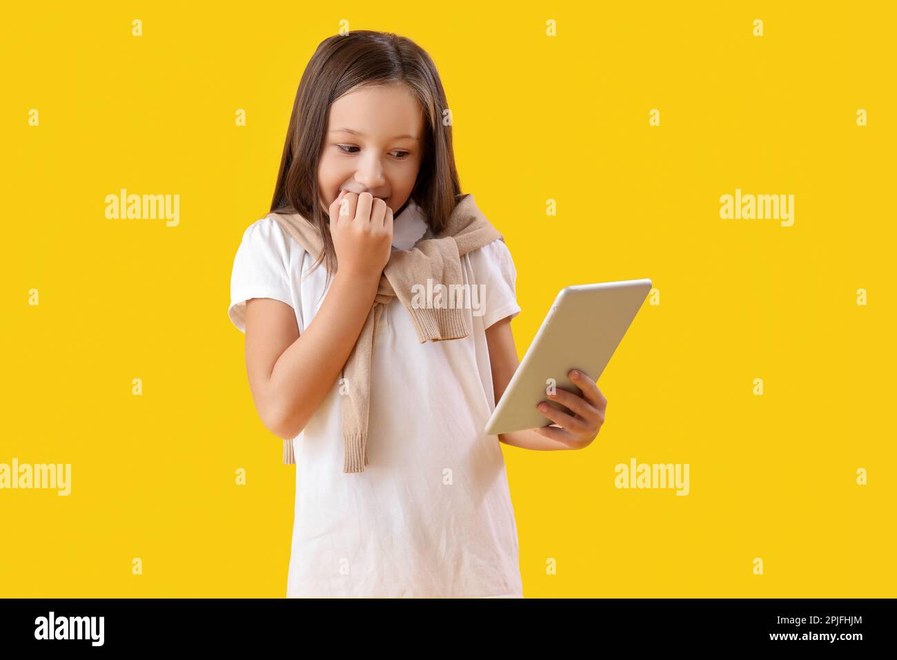 Little girl with tablet computer biting nails on yellow background ...