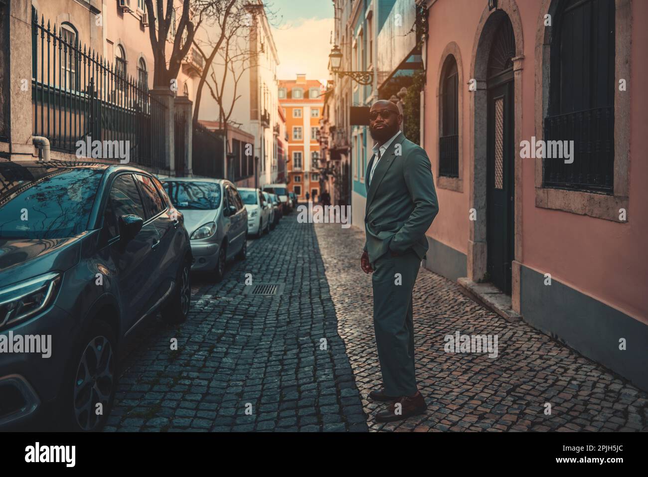 A man in a stylish grey-green suit and sunglasses stands on a narrow ...