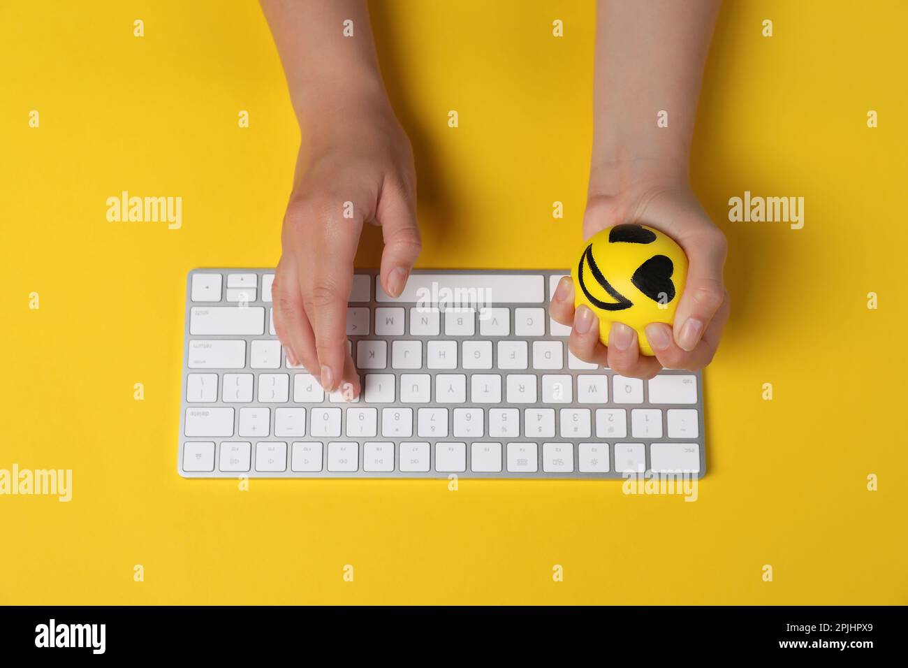 Woman squeezing antistress ball while typing on keyboard against yellow ...
