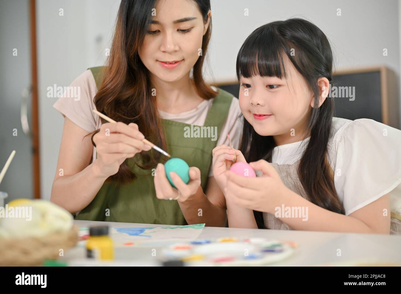 Caring and kind Asian mom teaching her little daughter to paint and color Easter eggs, spending ...