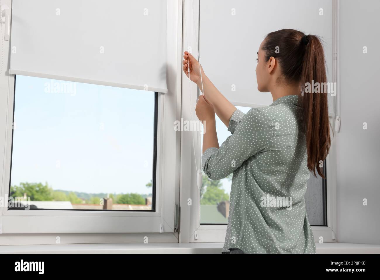 Woman opening white roller blind on window indoors Stock Photo - Alamy