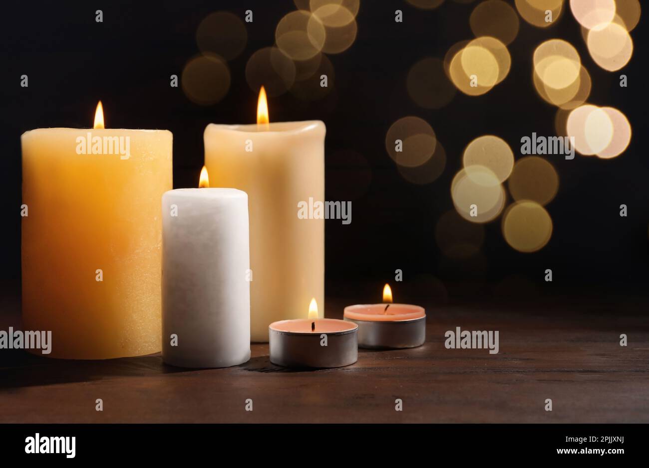 Different wax candles burning on table against dark background with blurred lights. Bokeh effect ...