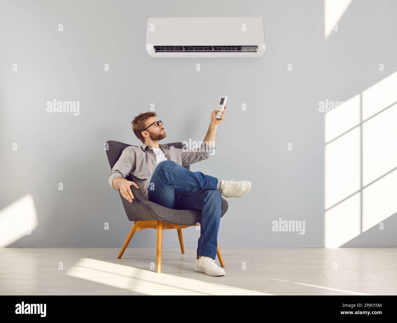 Young bearded man turning on air conditioner sitting on chair in a ...