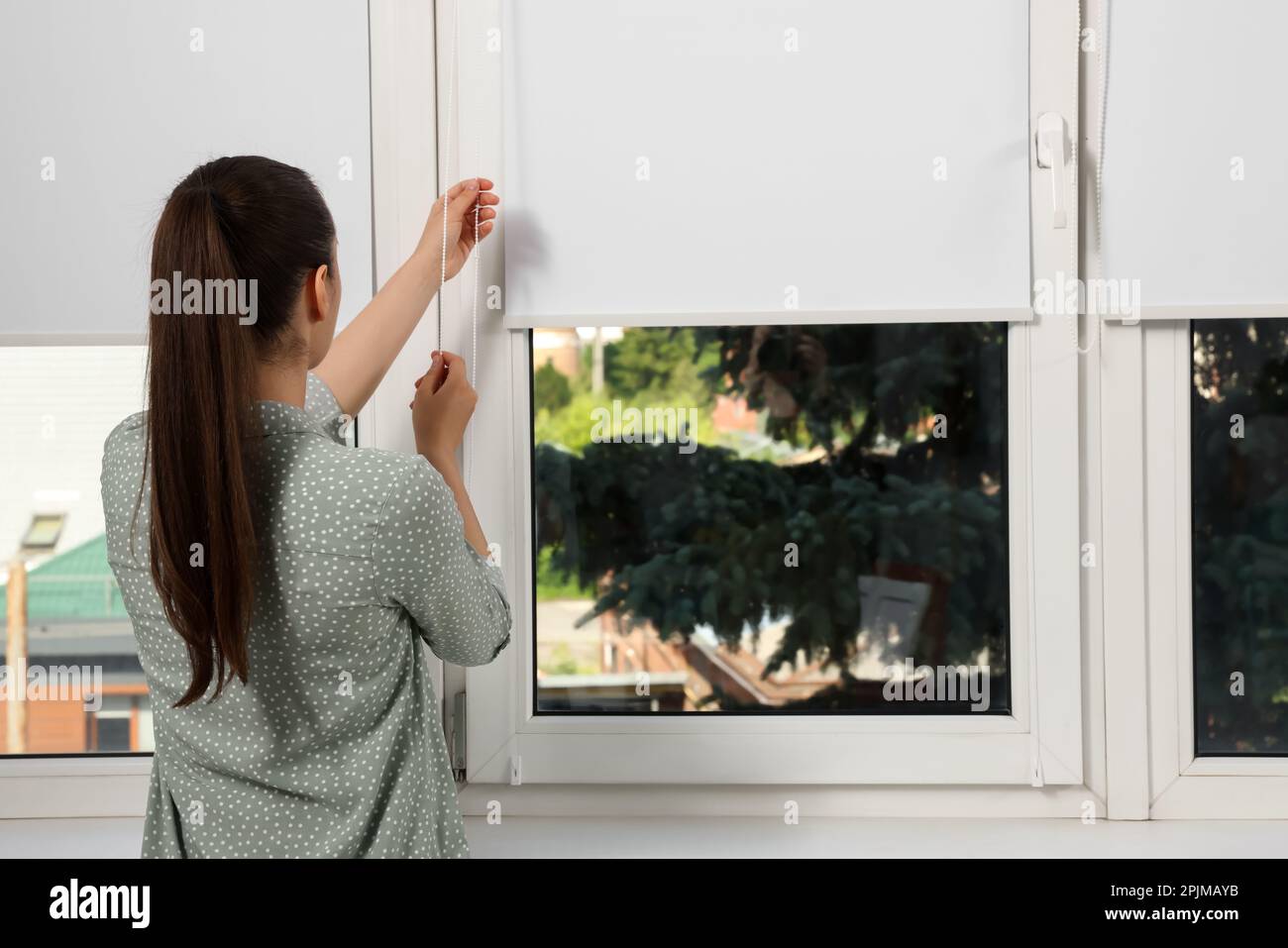 Woman opening white roller blind on window indoors, back view Stock ...