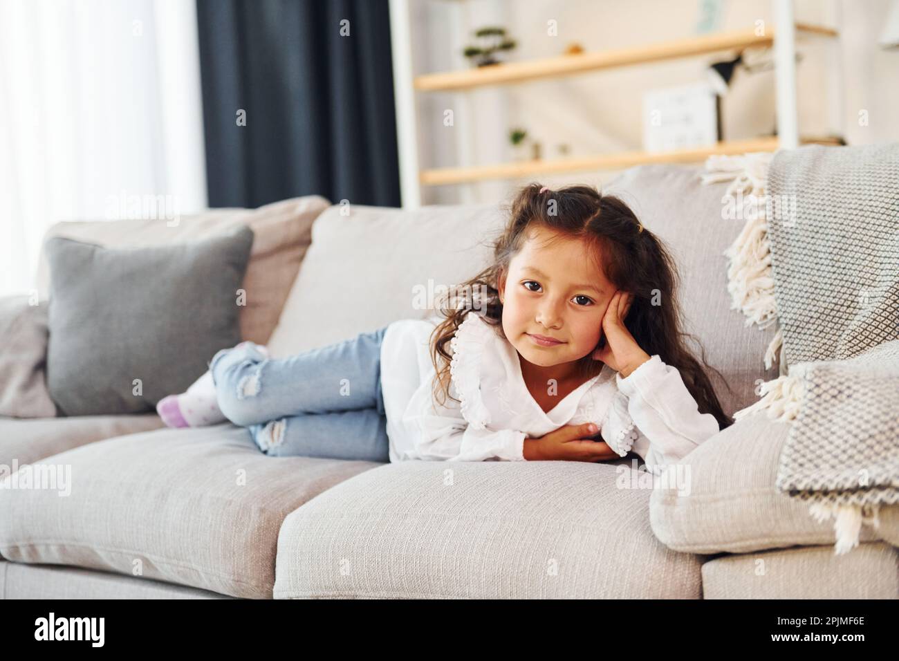 Laying down on the sofa. Little girl in casual clothes is at home alone ...