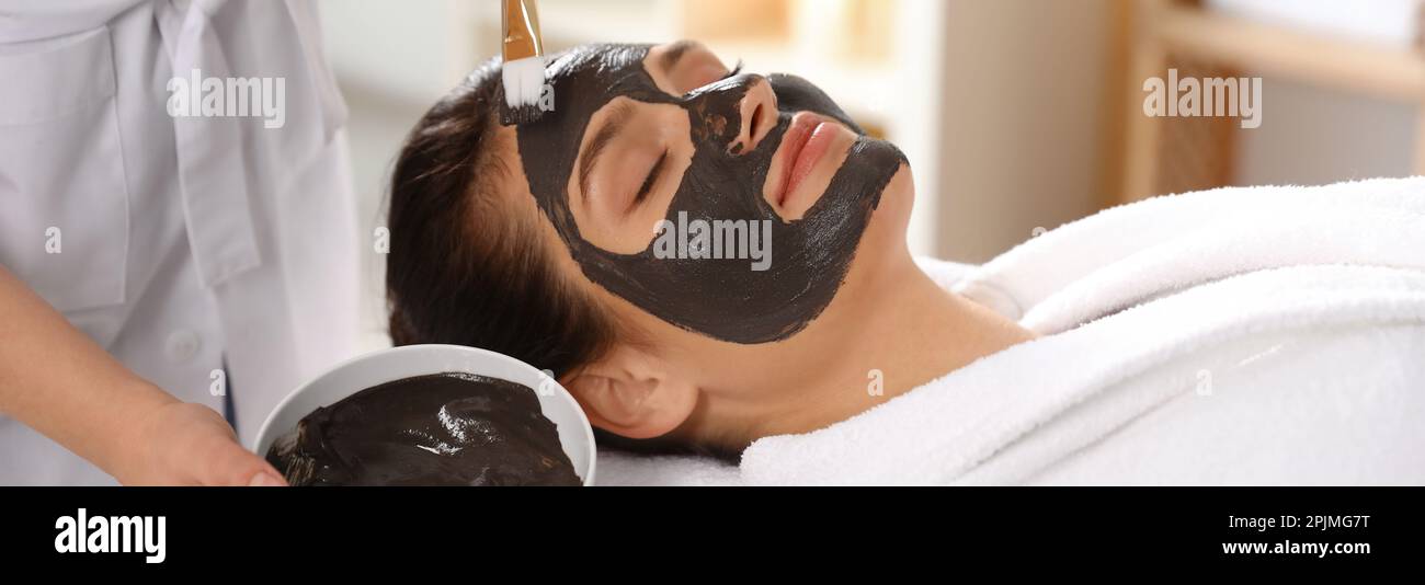 Cosmetologist applying black mask onto woman's face in spa salon ...