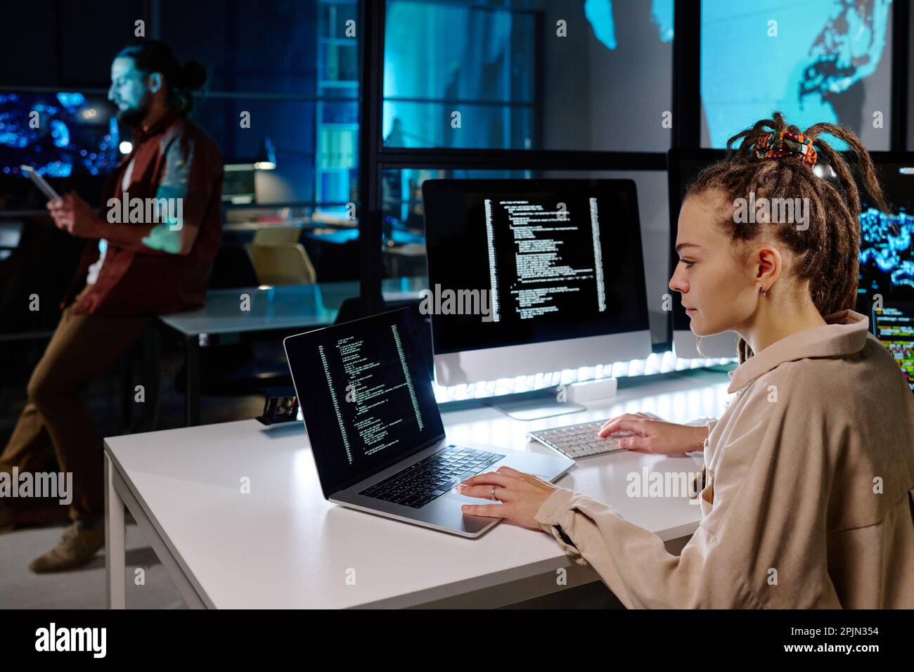 Serious female programmer with dreadlocks decoding data on laptop screen while sitting by desk ...