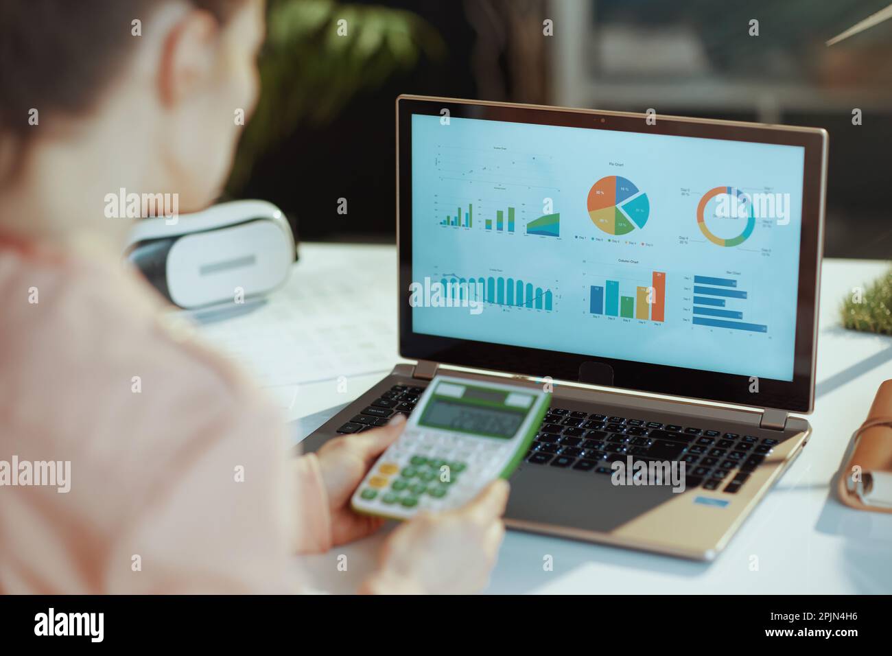 Seen from behind modern female with calculator and laptop working with graphs in modern office ...