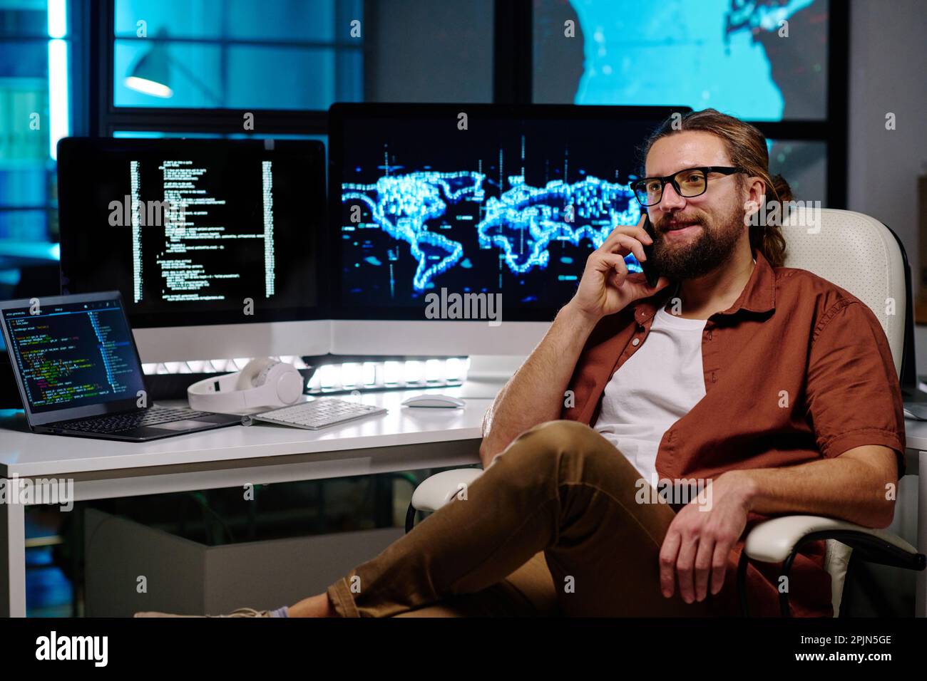 Young smiling IT manager speaking on mobile phone while sitting by ...