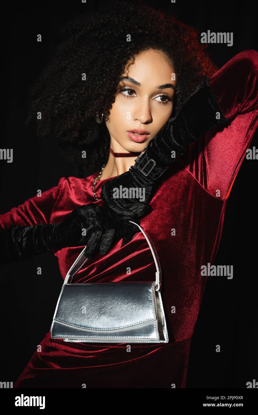 Trendy african american model in velvet dress and gloves holding silver ...