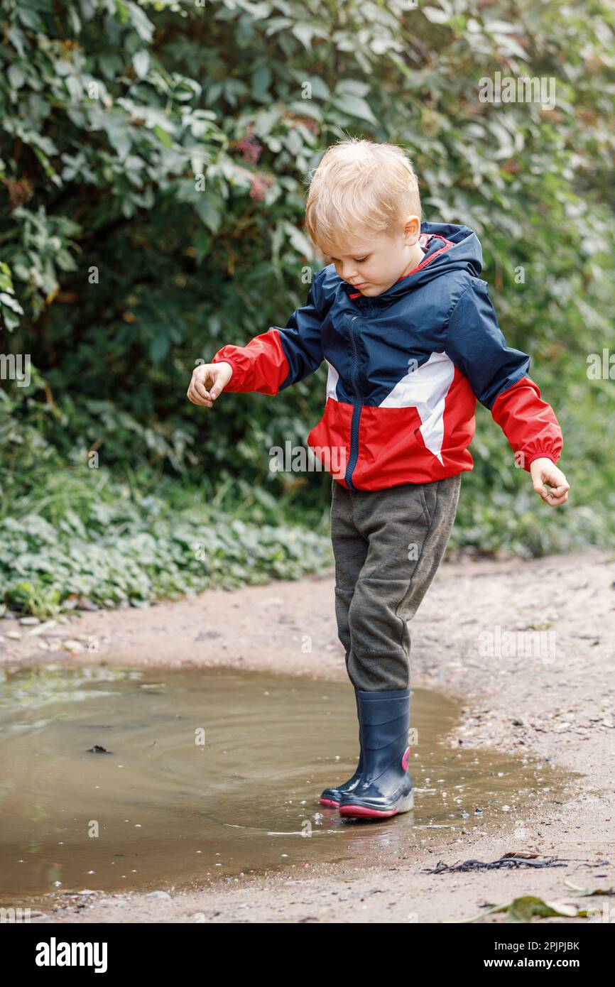A young blond boy in a blue and red raincoat and rain boots happily go ...