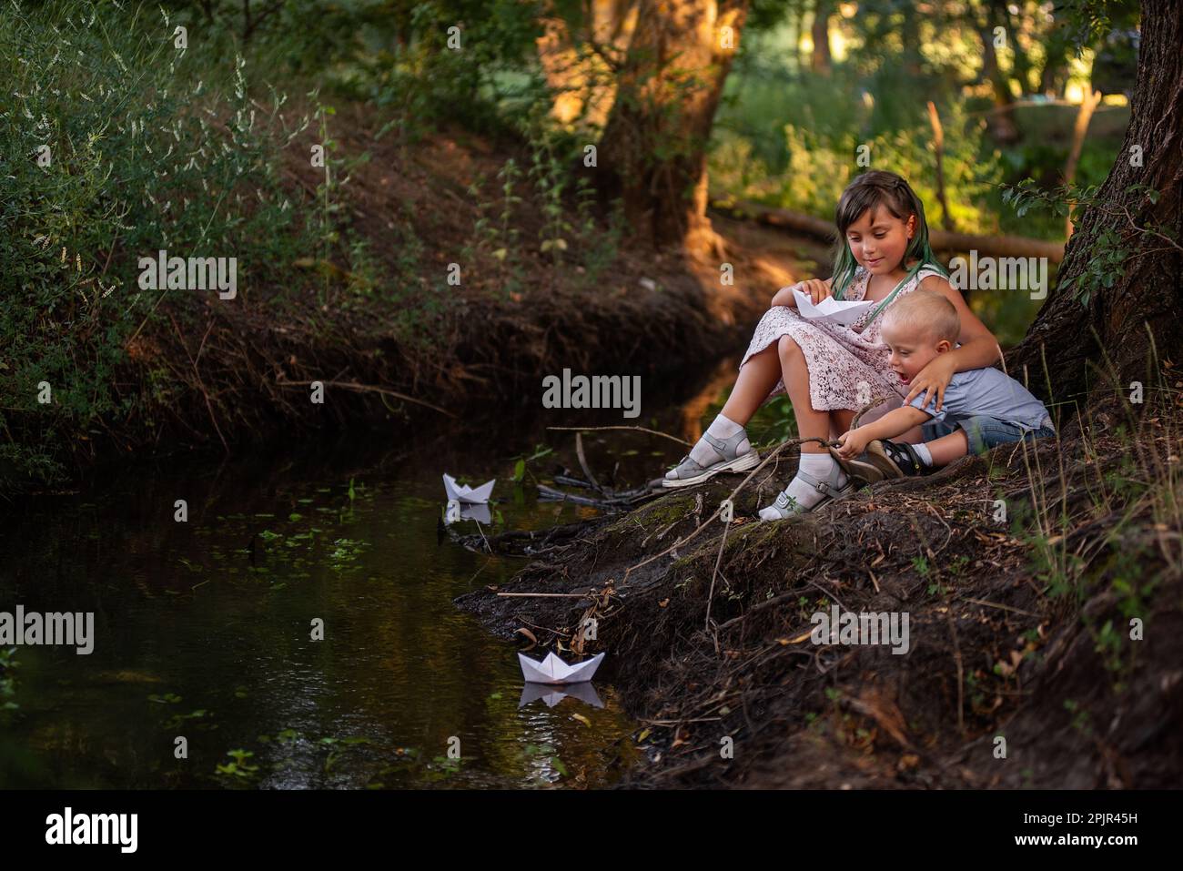 Girl and boy are sitting on the bank of river, launching white paper ...