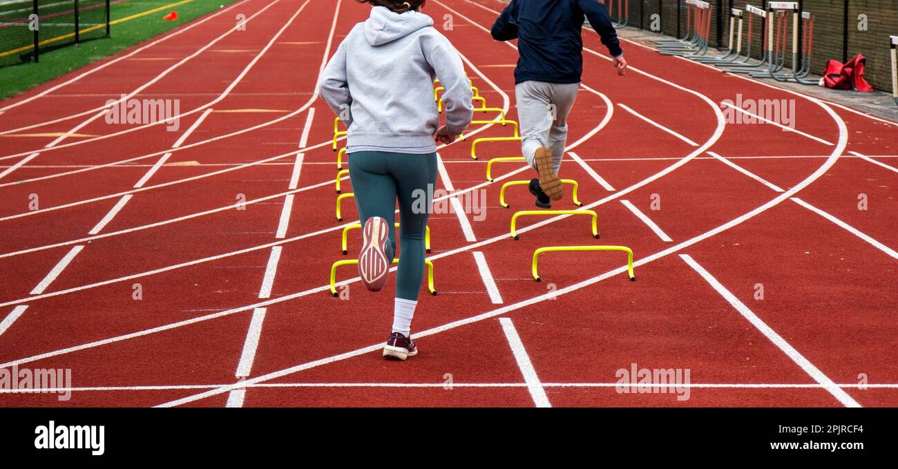 Rear view of two runners running over six inch yellow mini hurdles in lanes on a track at ...