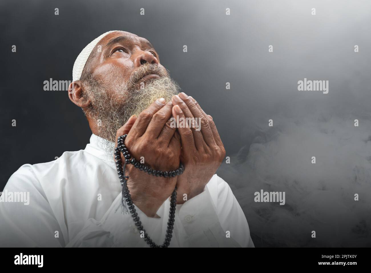 Muslim man with a beard wearing a white cap praying with prayer beads ...