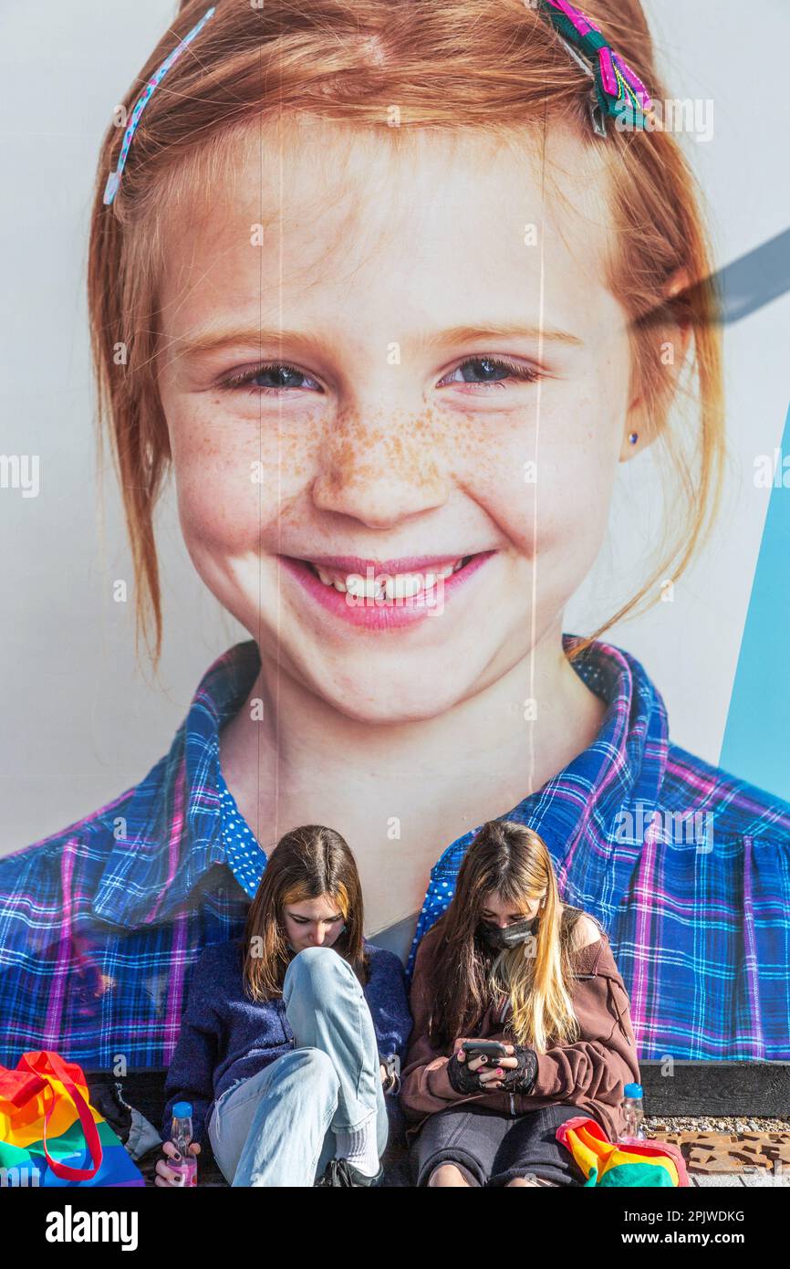 Two young women seated on the ground, at the foot of a large poster ...