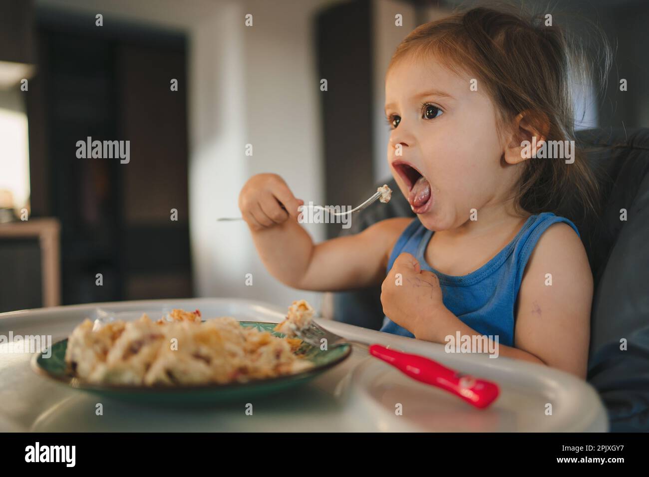 Toddler baby girl learnsing to eat herself with a fork while sitting in ...