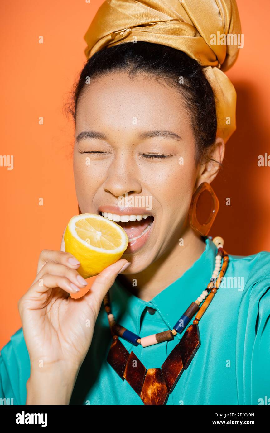 Trendy african american woman biting fresh lemon on orange background ...