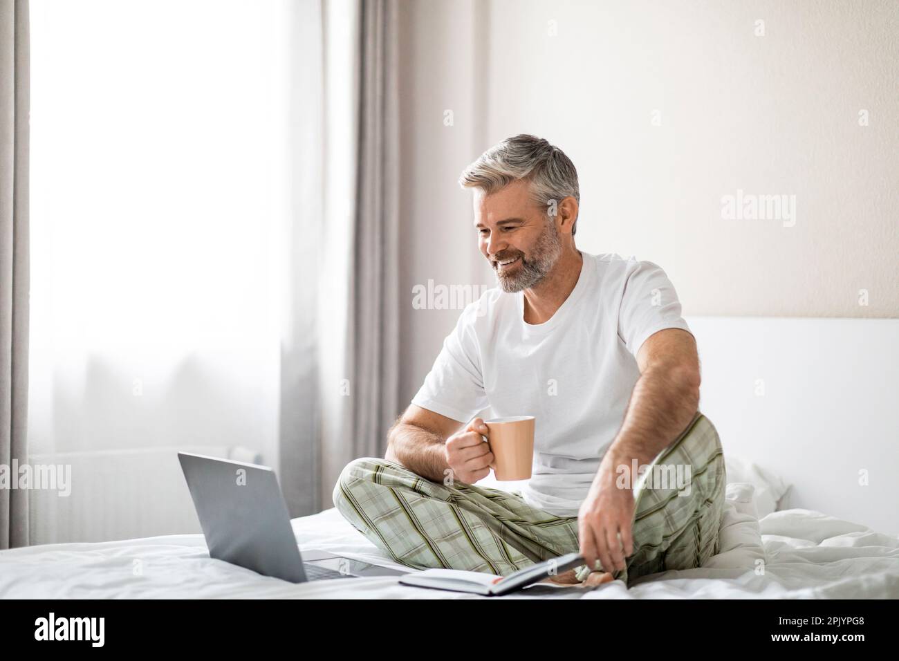 Middle aged man sitting on bed with laptop and notepad Stock Photo - Alamy