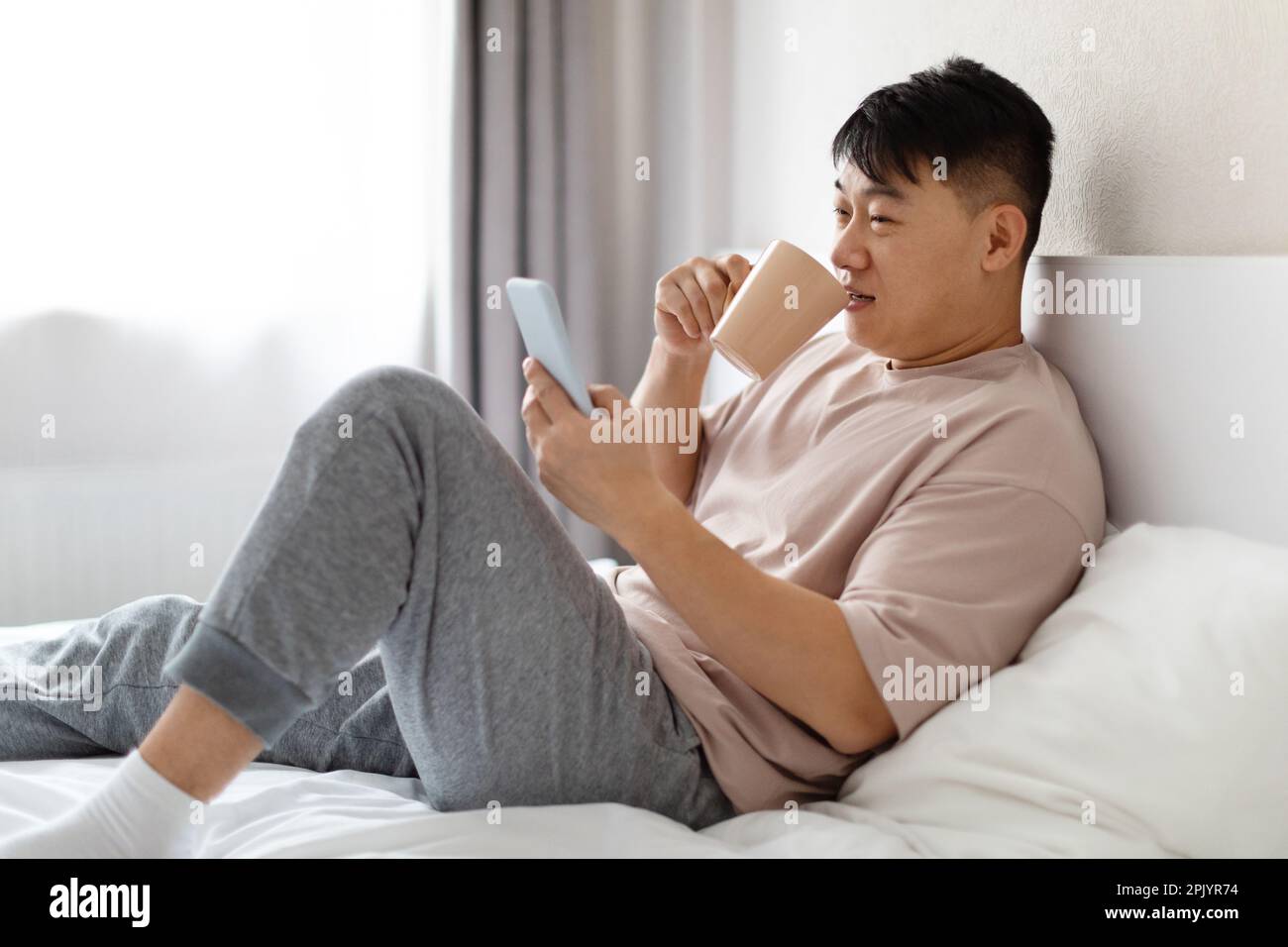 Relaxed chinese man chilling in bed, using phone, drinking coffee Stock ...