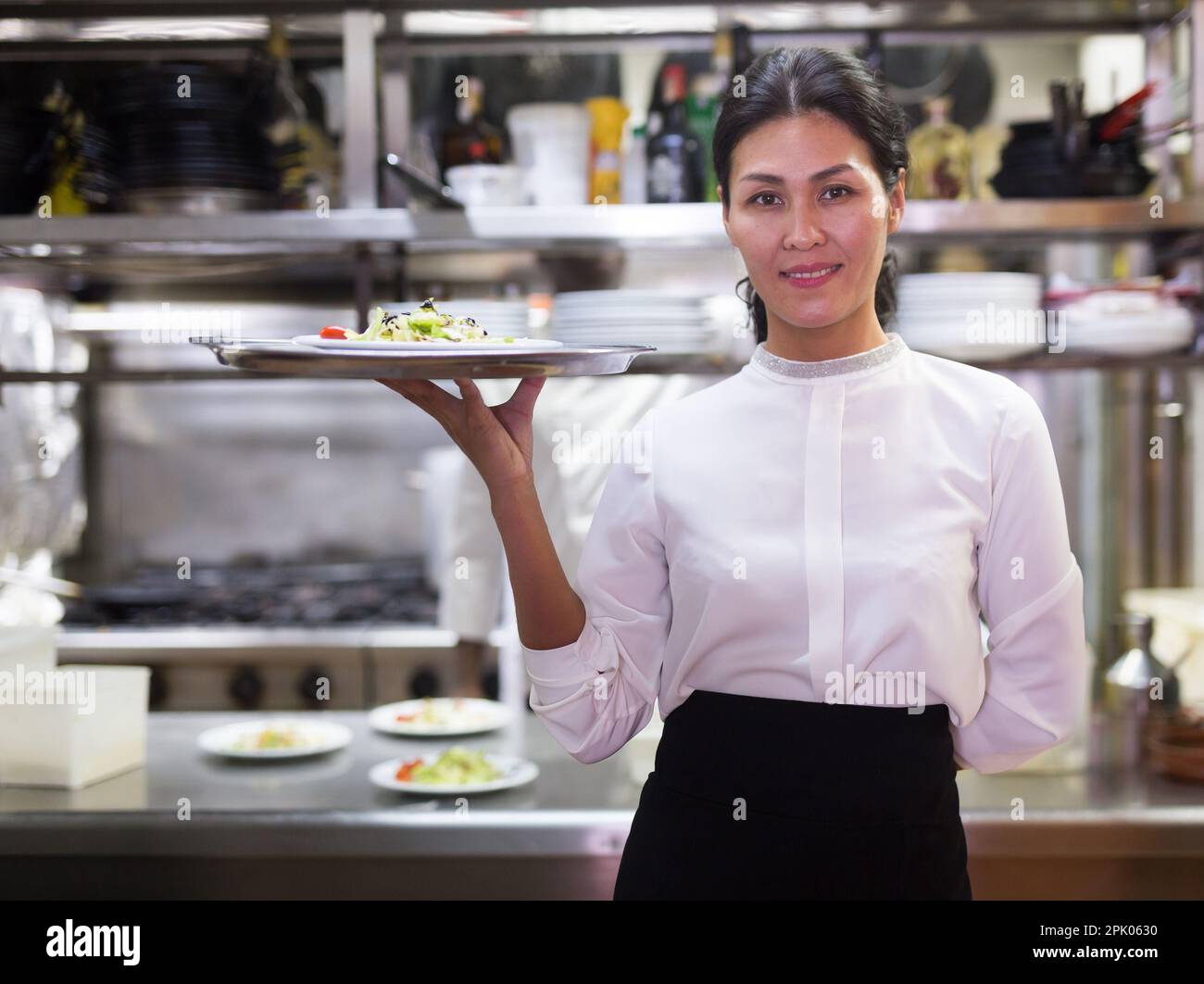 Successful waitress standing in restaurant kitchen with ordered meals ...