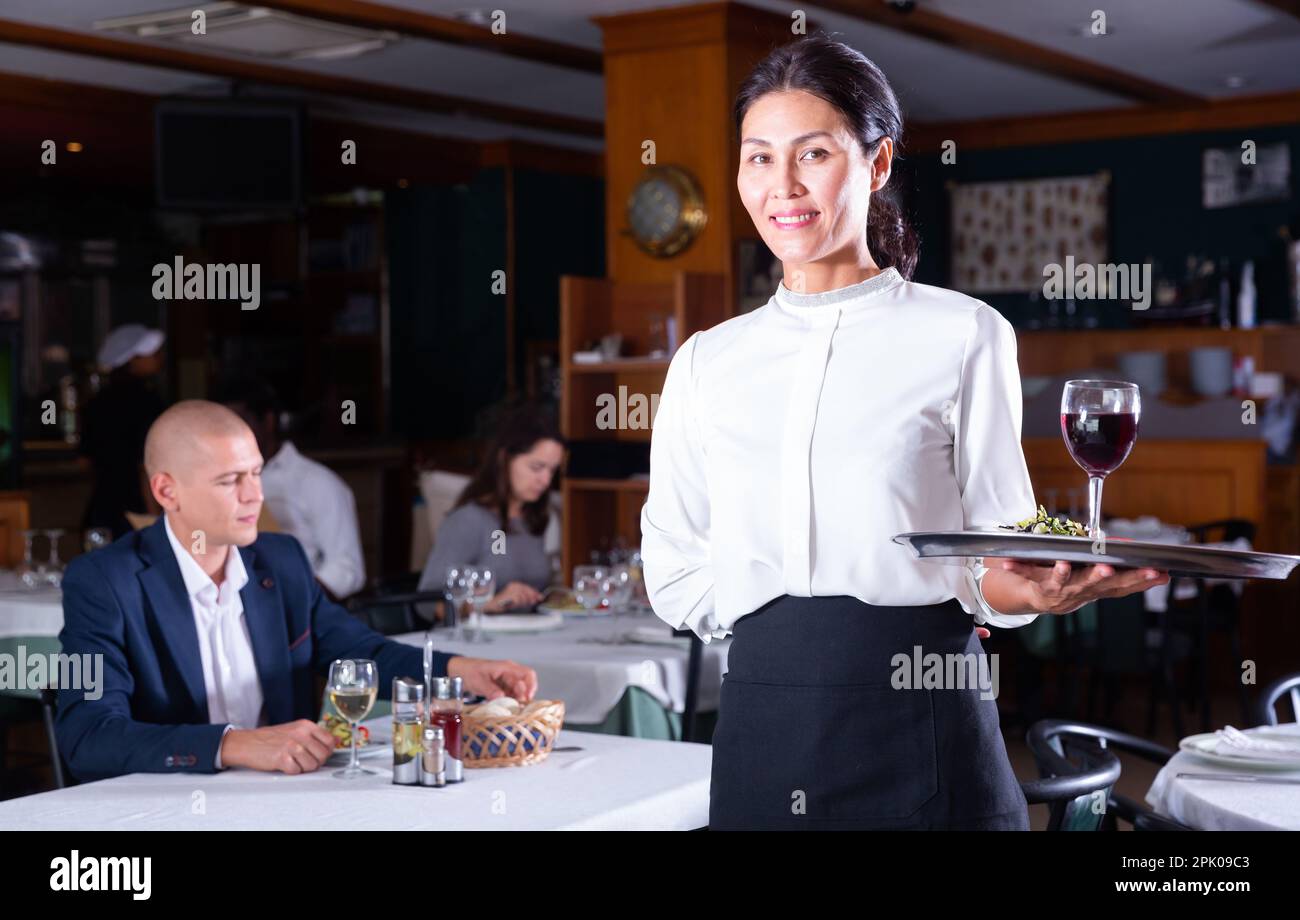 Hospitable waitress with serving tray meeting guests in restaurant ...
