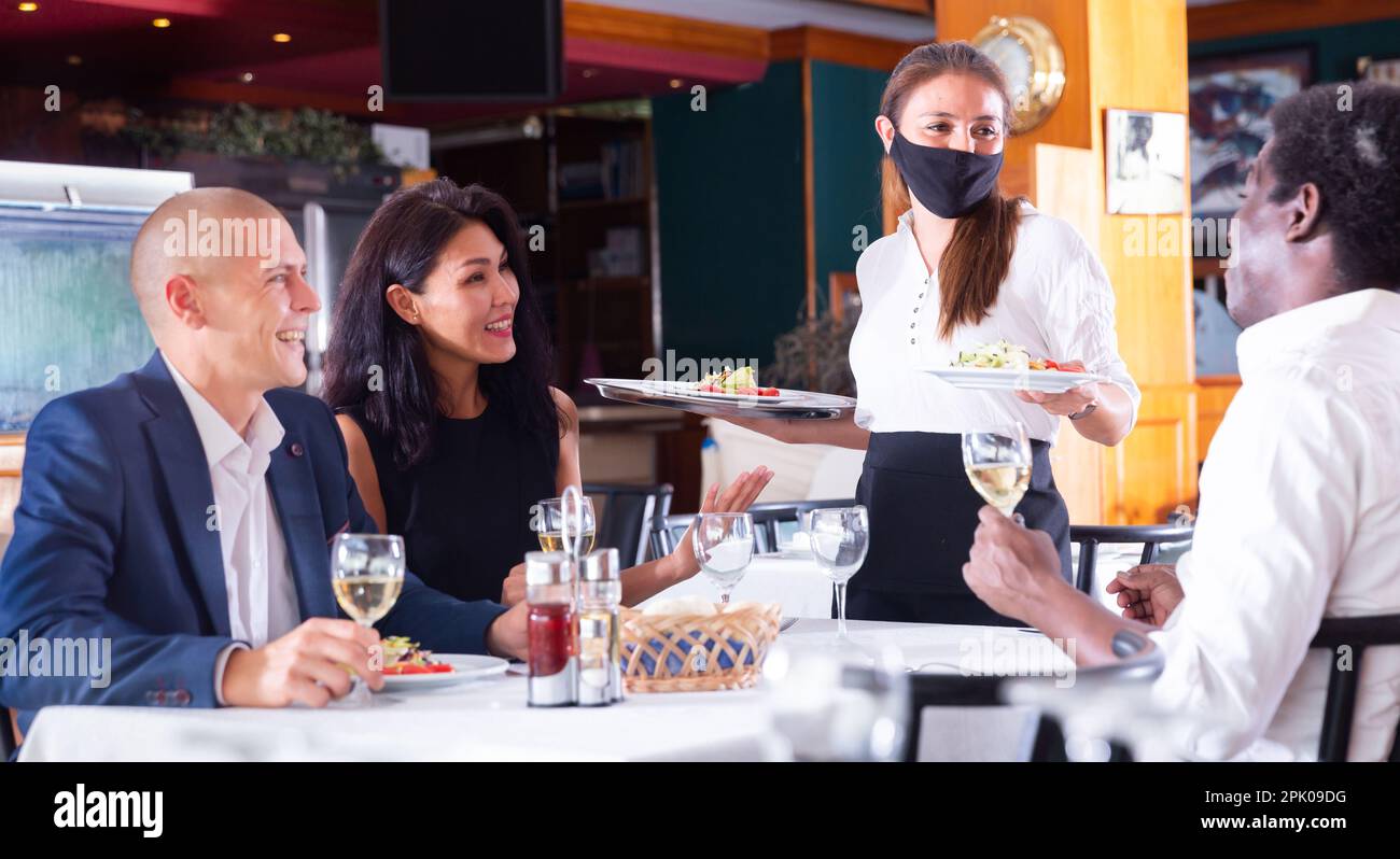smiling female waiter bringing order to group of friens in restaurante ...