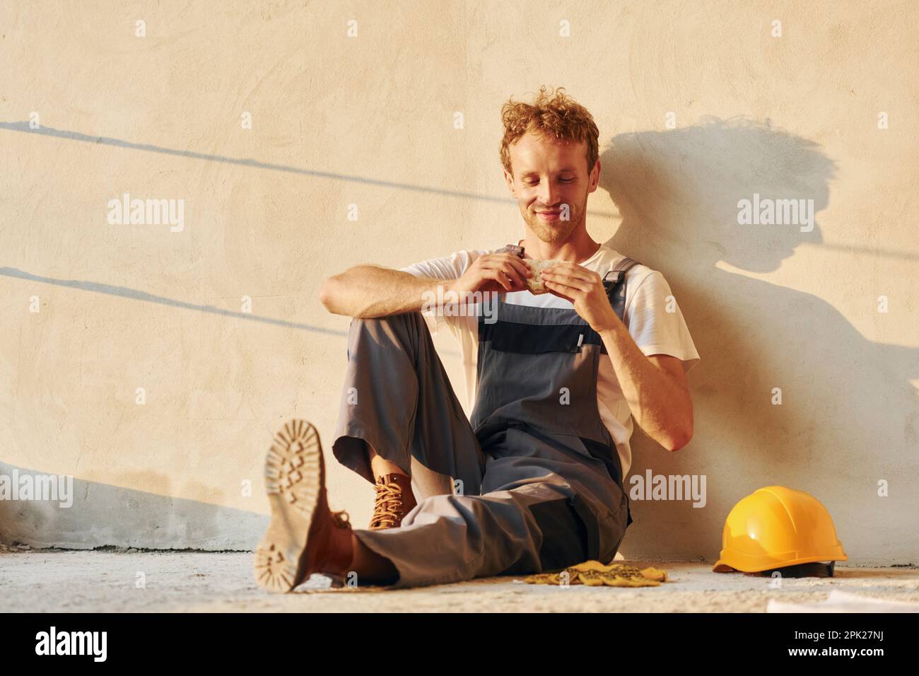 Eating food. Young man working in uniform at construction at daytime ...