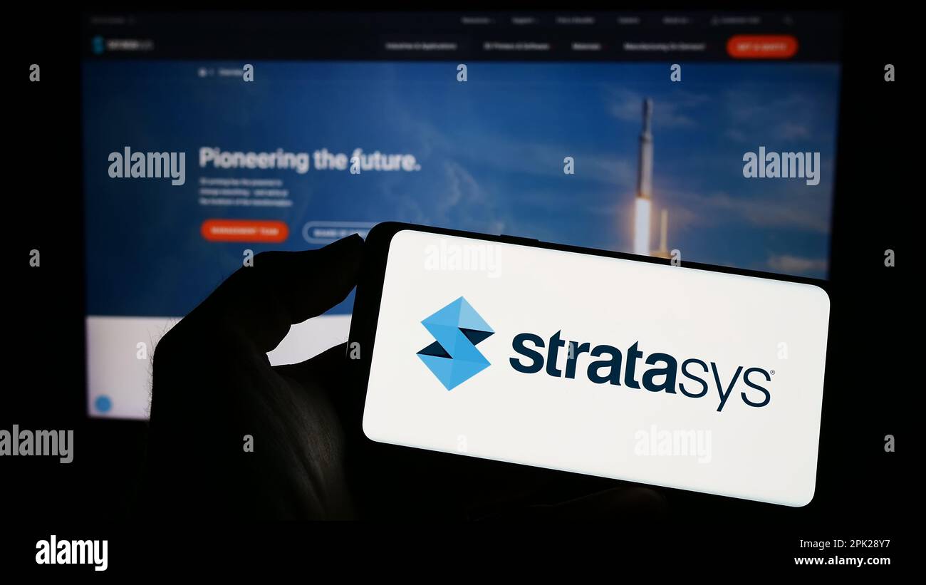 Person holding mobile phone with logo of 3D printing company Stratasys Ltd. on screen in front ...