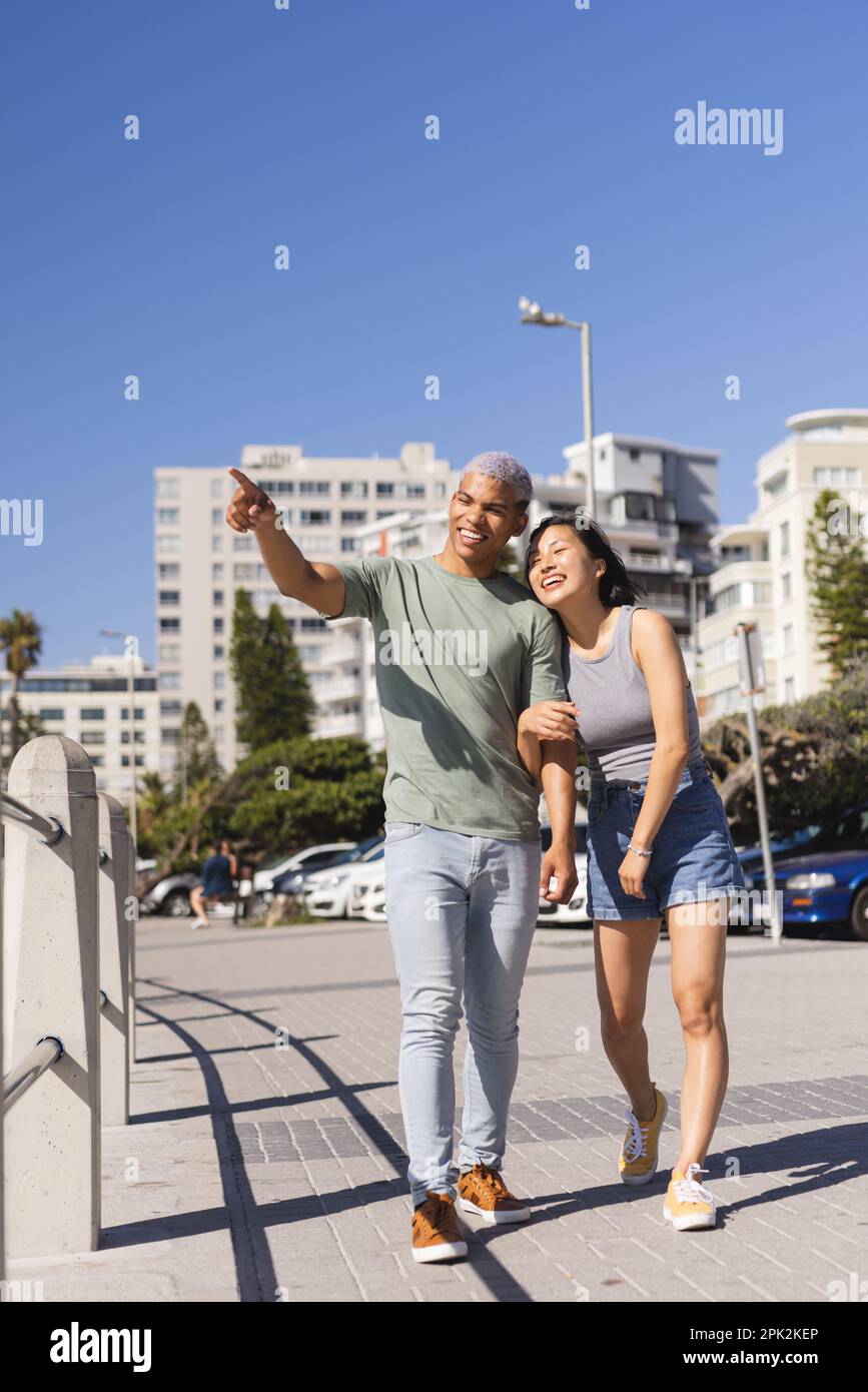 Happy diverse couple pointing and walking along promenade by the sea ...