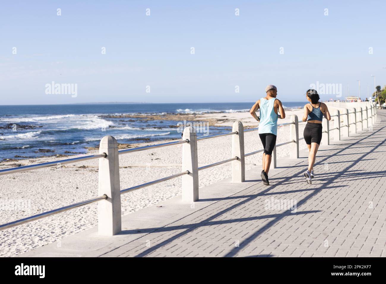 Diverse couple wearing sportswear and running on promenade by the sea ...